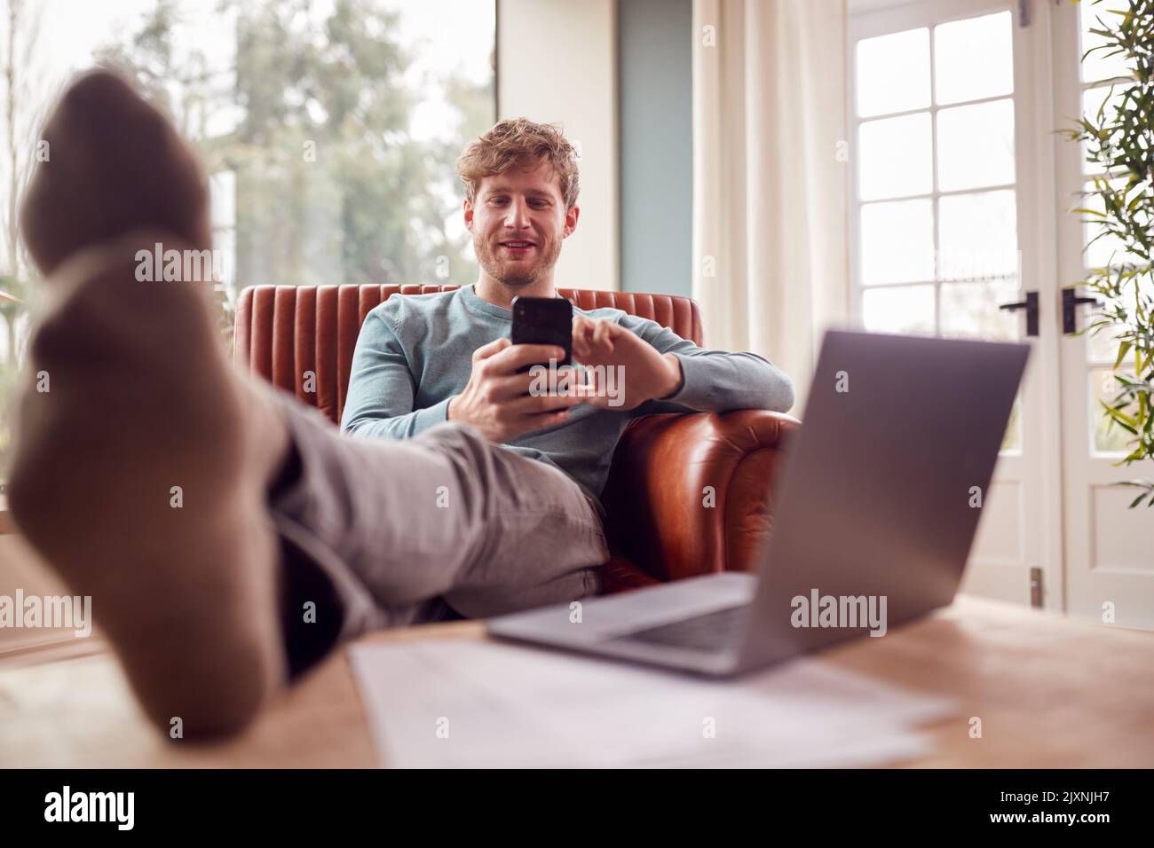 Man Relaxing In Armchair With Feet Up At Home Using Laptop And Mobile ...