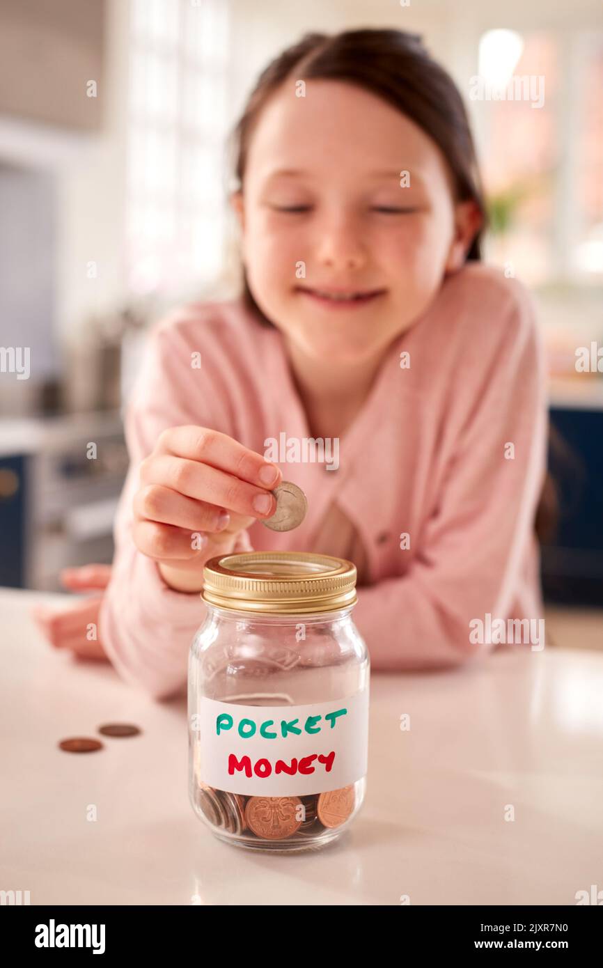 Girl Saving Money Into Jar Labelled Pocket Money On Kitchen Counter At ...