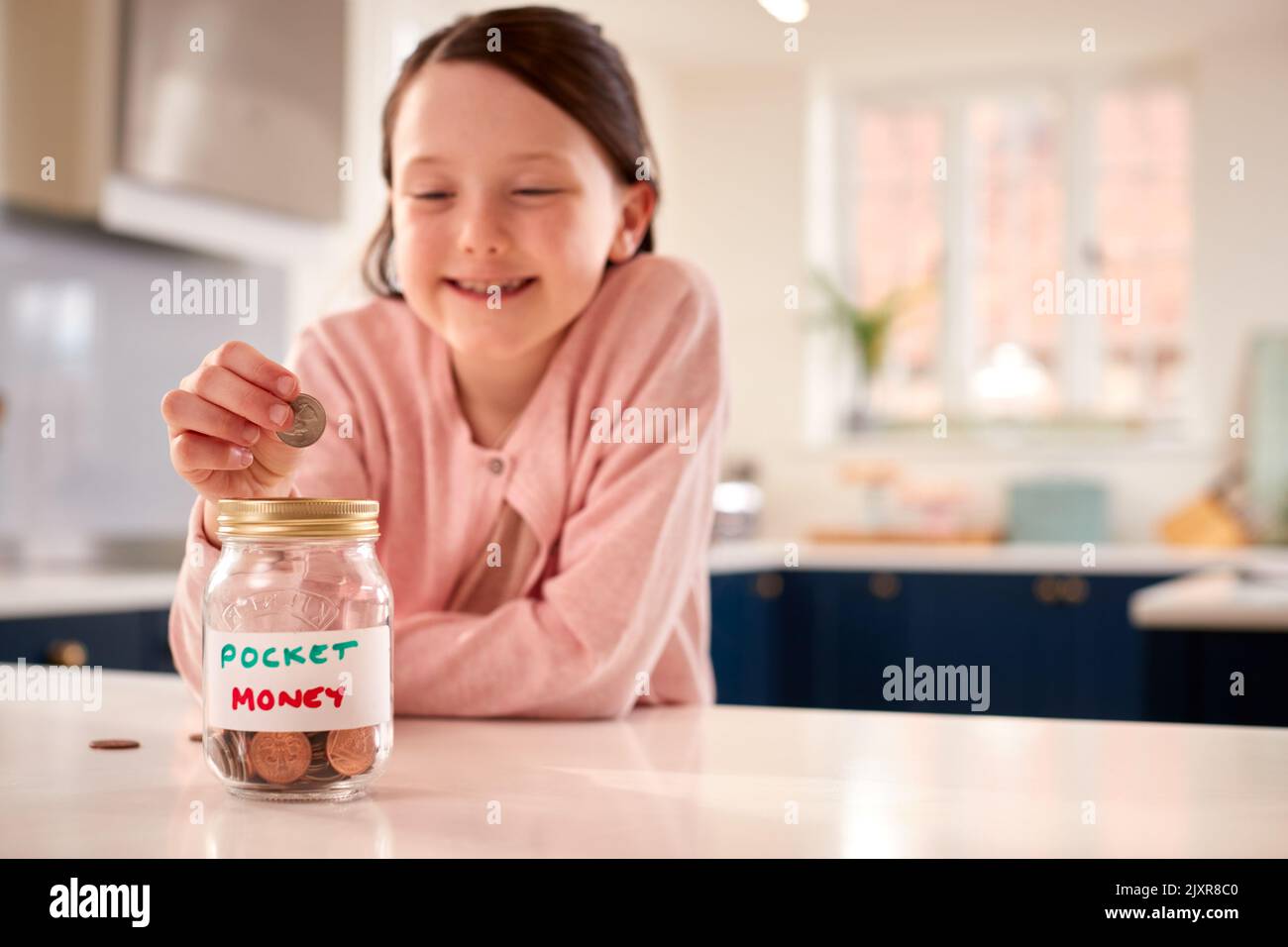 Girl Saving Money Into Jar Labelled Pocket Money On Kitchen Counter At ...