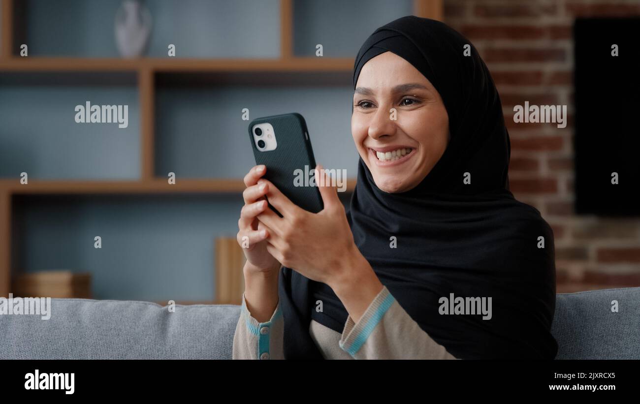 Smiling Arab Muslim Islamic 30s woman user in headscarf black hijab using phone at home sitting ...