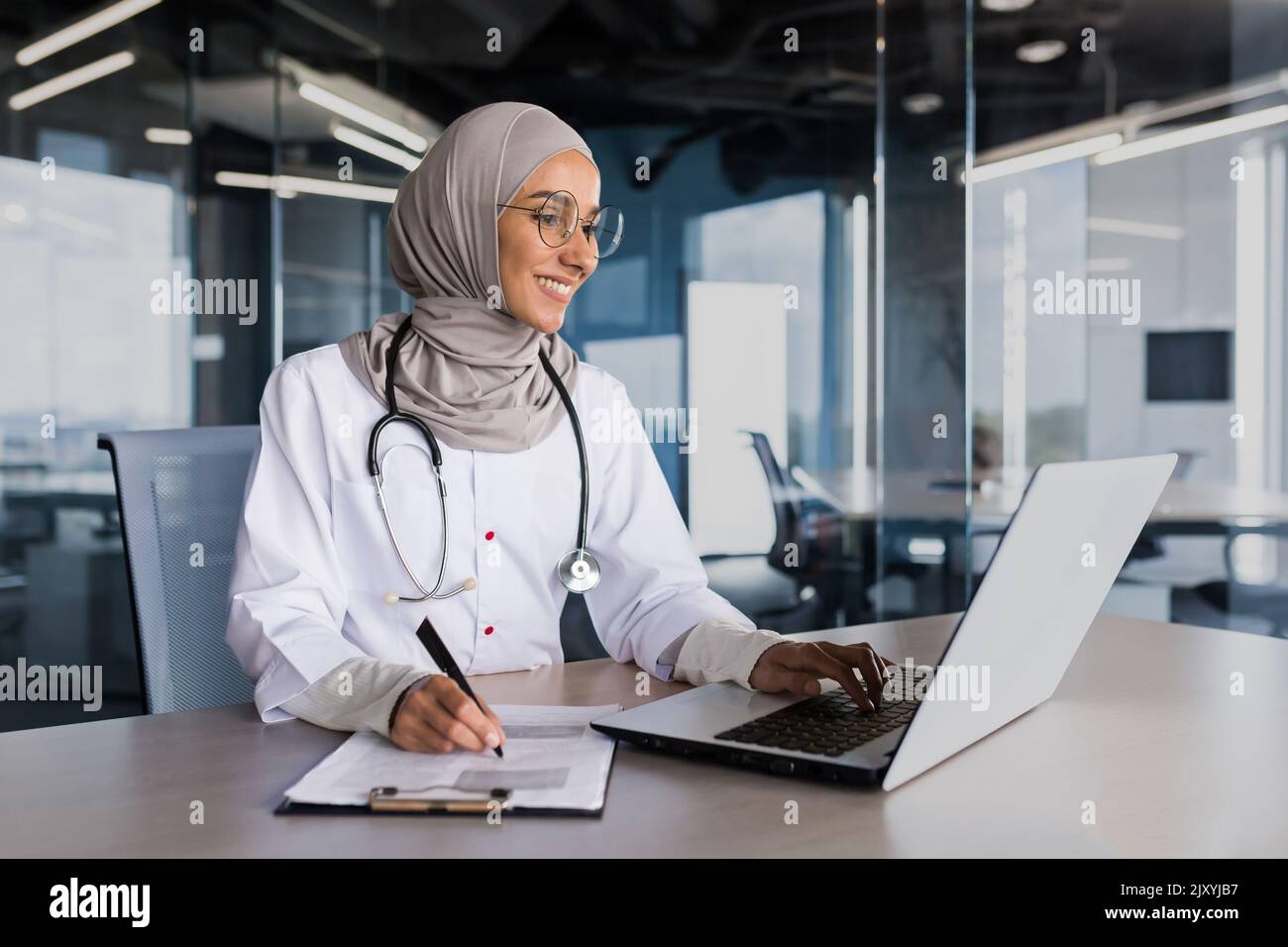 Arab muslim woman in hijab doctor working in modern clinic office with laptop, doctor on paper ...