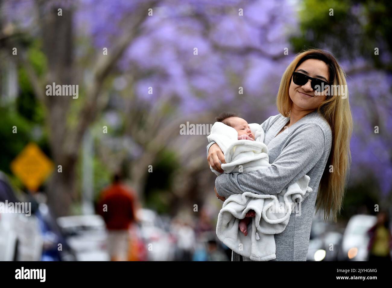 A woman holding a baby poses for a photo amongst jacaranda trees at ...