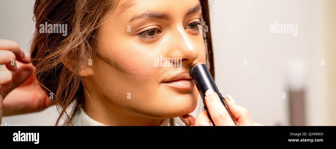 Beautiful young brunette woman receiving makeup with stick concealer on ...
