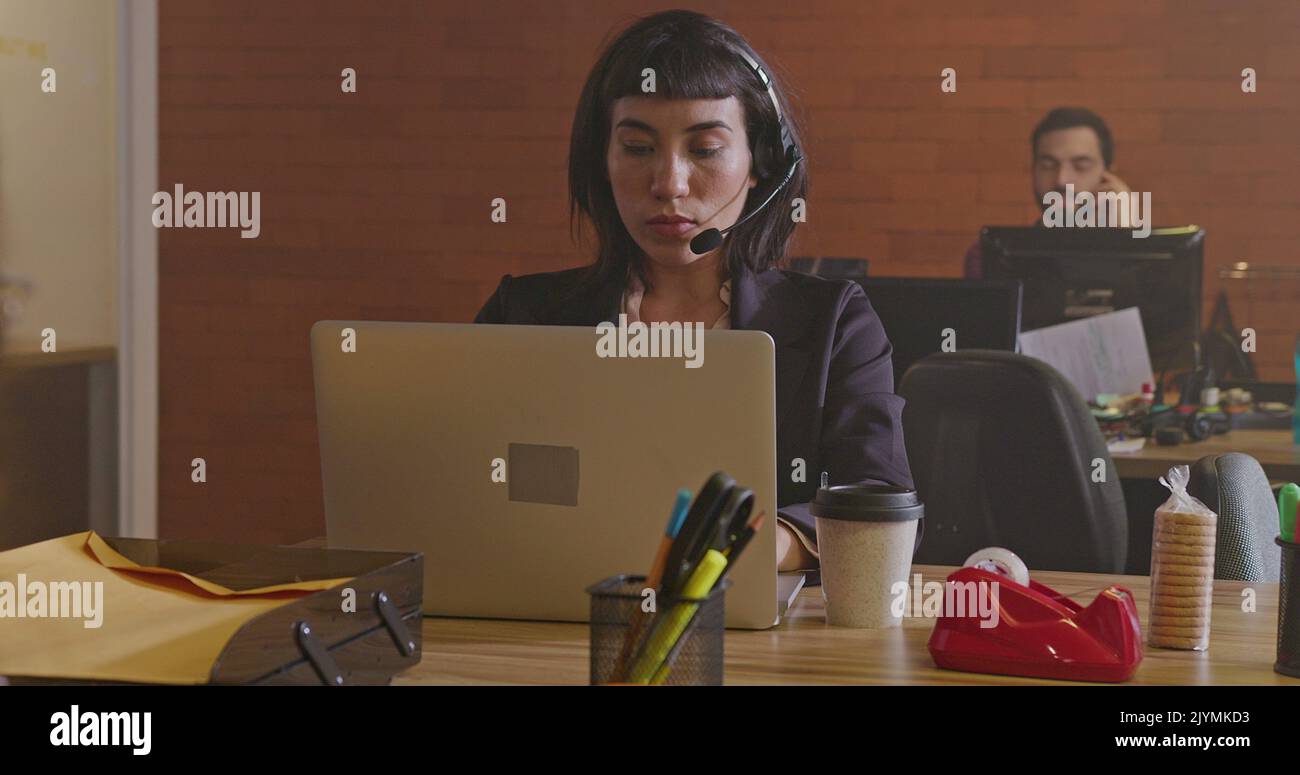A hispanic young female employee wearing headset sitting in front of ...