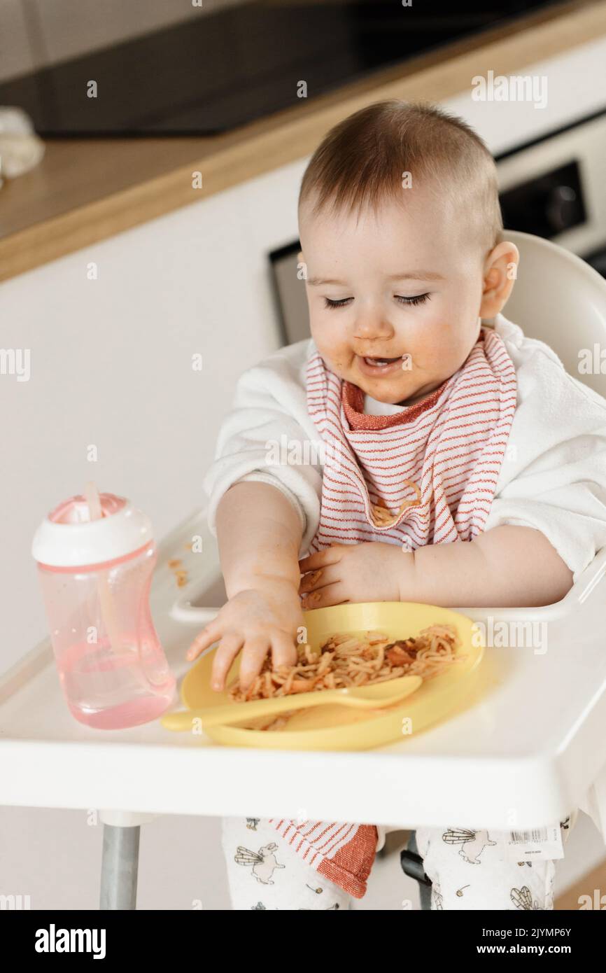 A young baby sitting in a high chair feeding herself spaghetti with a ...