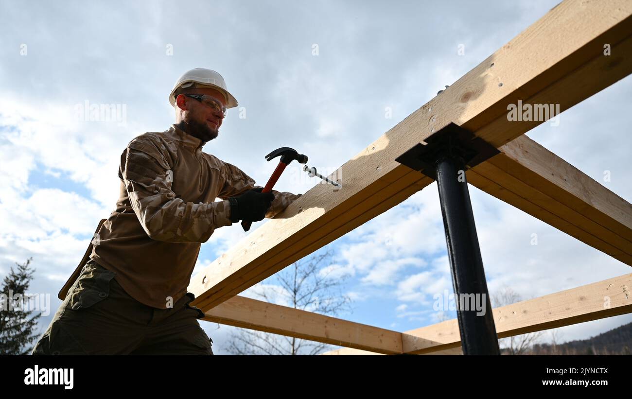 Man worker building wooden frame house on pile foundation. Carpenter hammering bolt into wooden ...