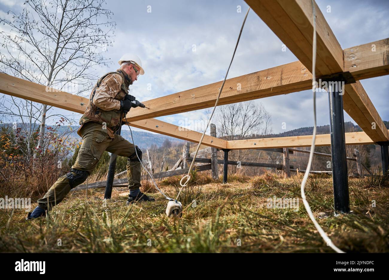 Man building wooden frame house on pile foundation. Side view of male worker drilling hole by ...