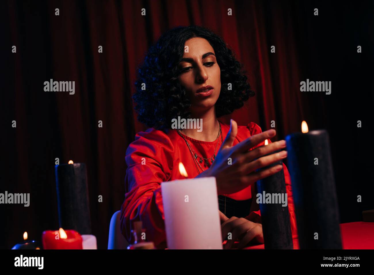 young fortune teller sitting near burning candles on dark background ...