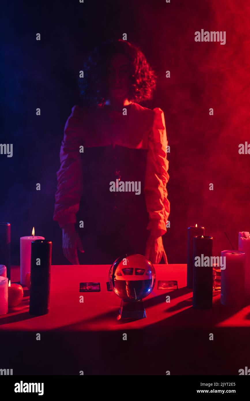 young fortune teller standing near red table with candles and crystal ...