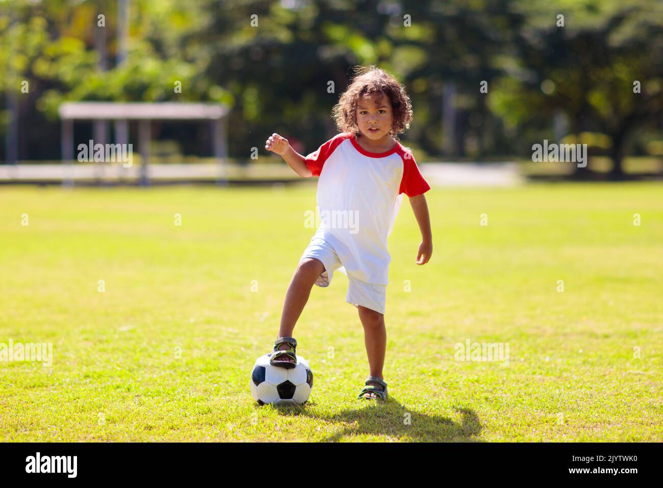 Cute curly little boy playing football. Kids play on outdoor pitch ...