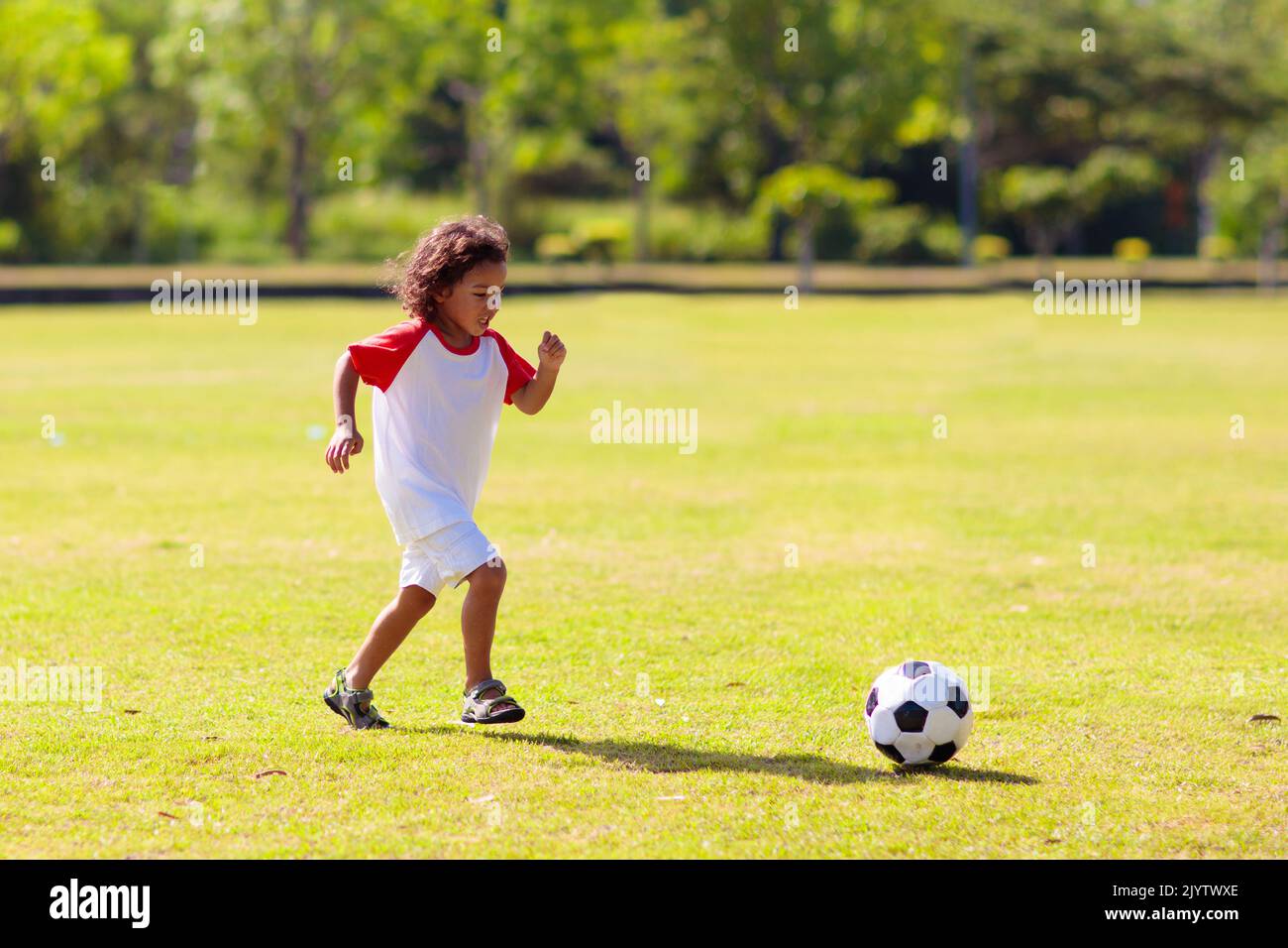 Cute curly little boy playing football. Kids play on outdoor pitch ...