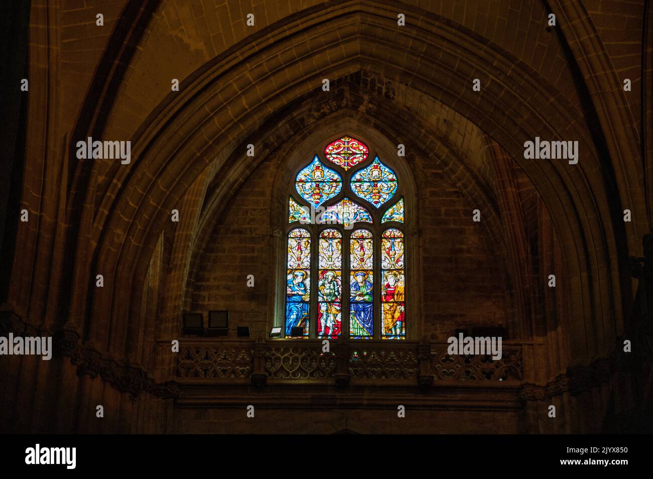 Colorful stained glass window set within a Gothic arch inside Seville Cathedral, illuminating ...