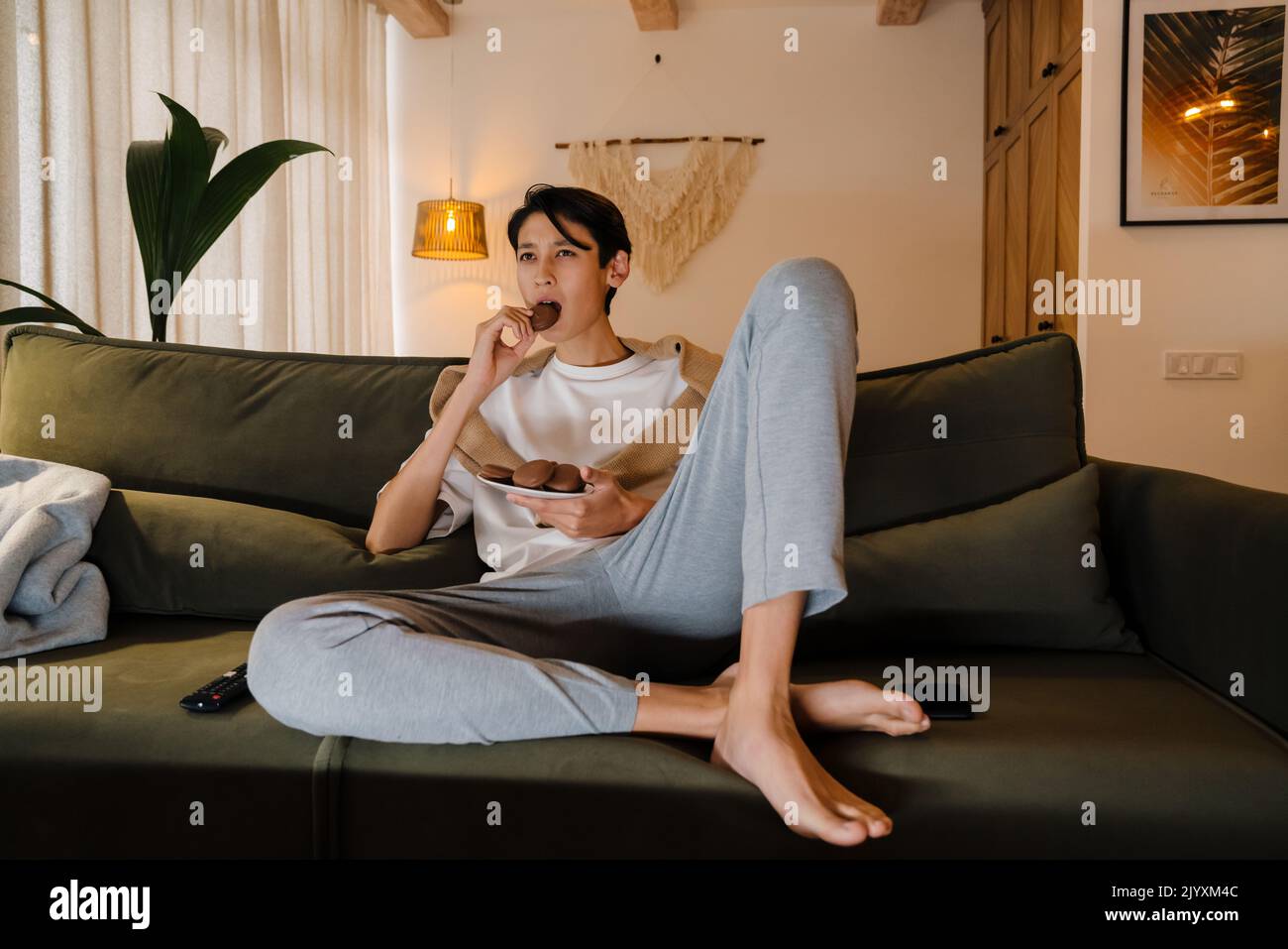 Asian boy eating cakes and watching tv while sitting on sofa at home ...