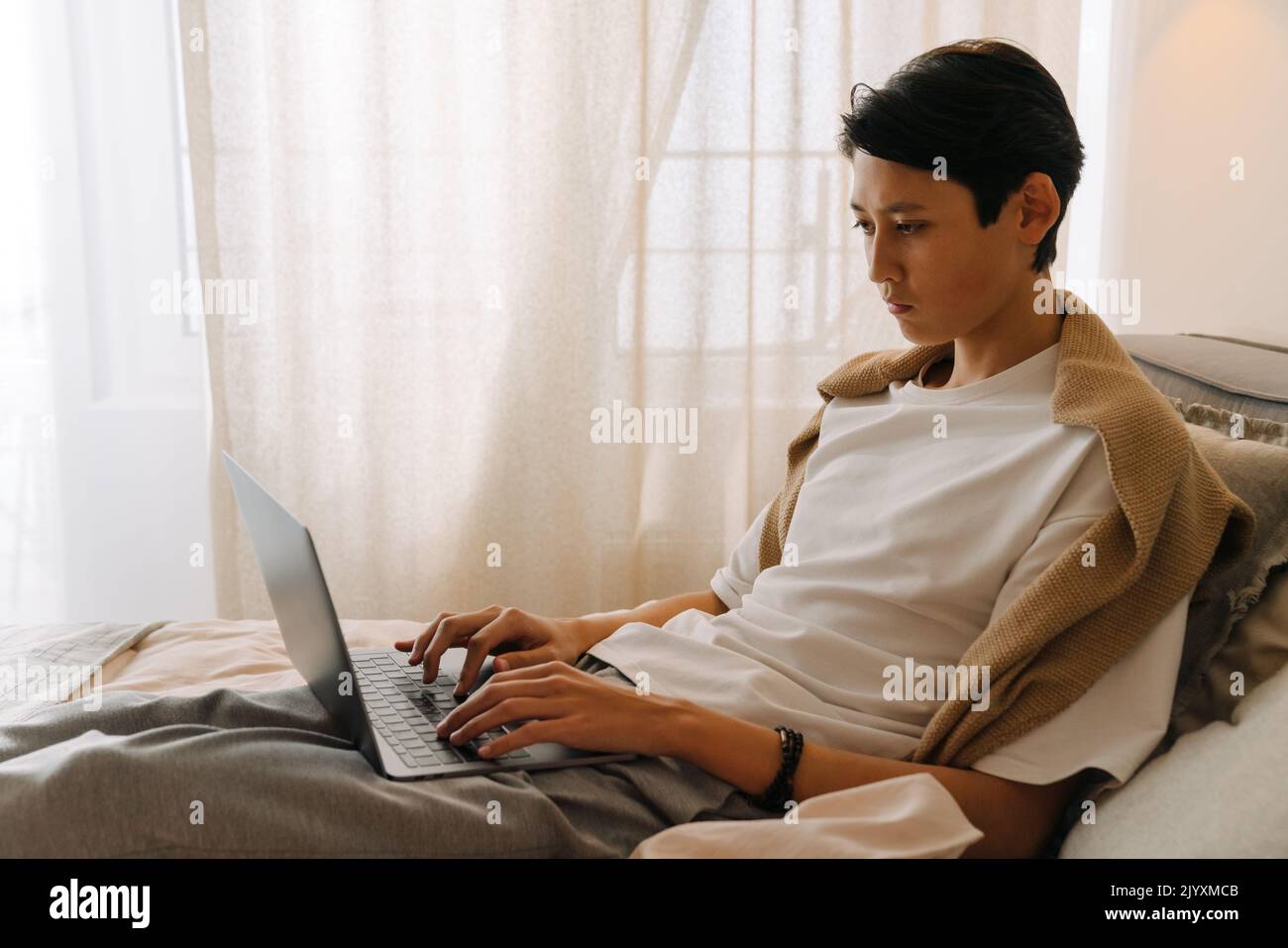 Asian boy working with laptop while resting on bed at home Stock Photo ...