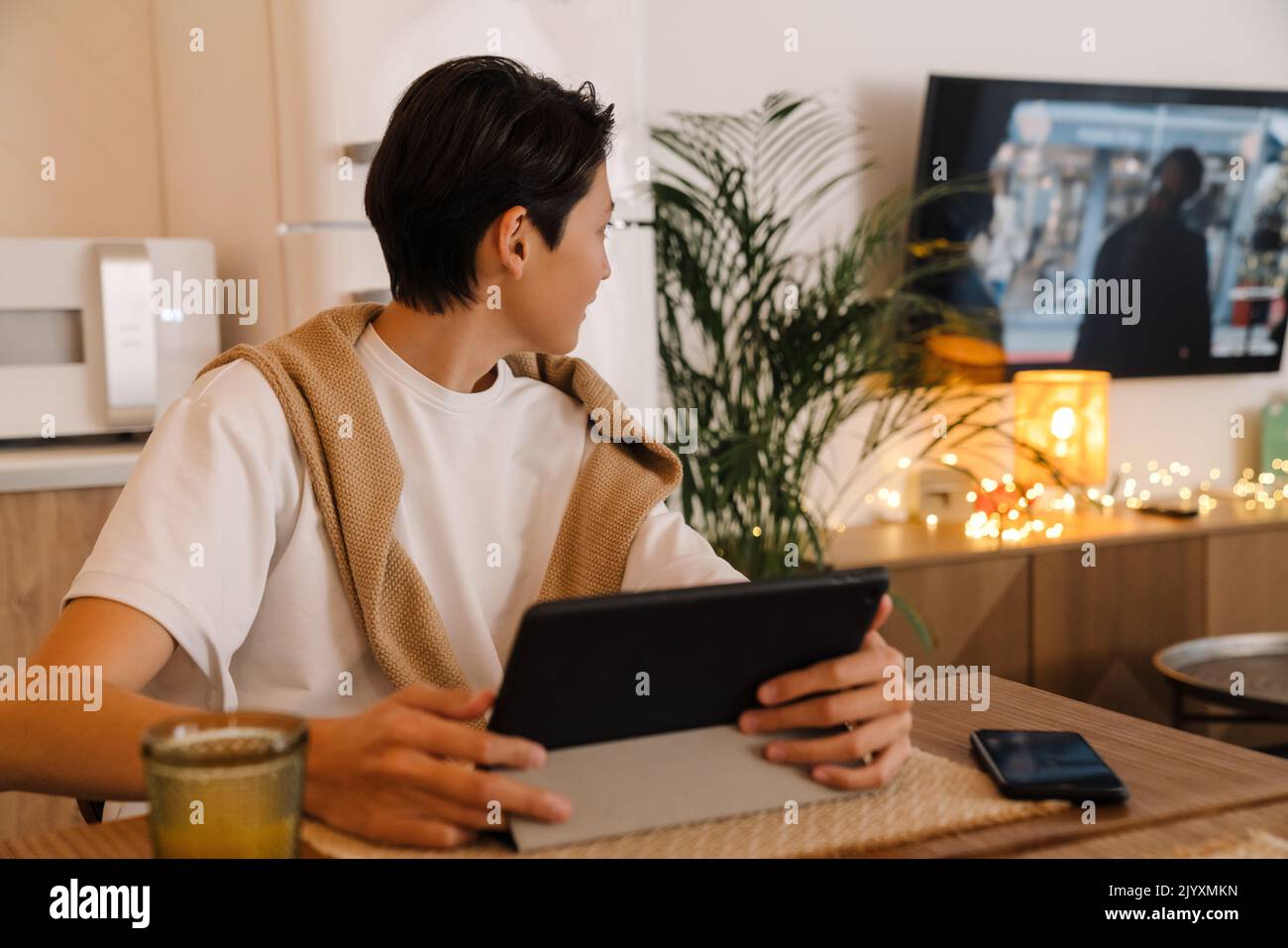 Asian boy using tablet computer while watching tv in kitchen at home ...