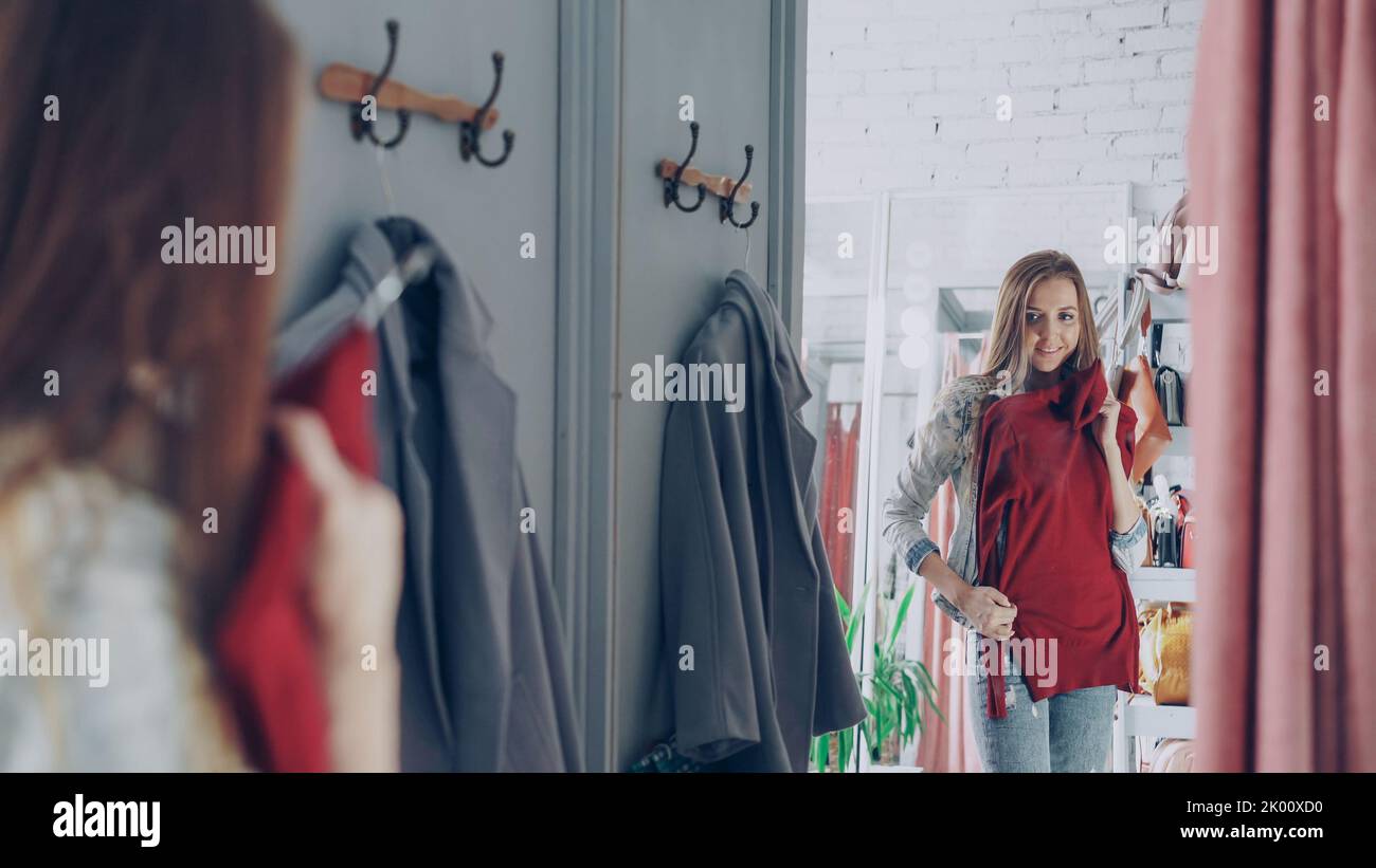 Mirror shot of young lady choosing clothes in fitting room. Girl is ...