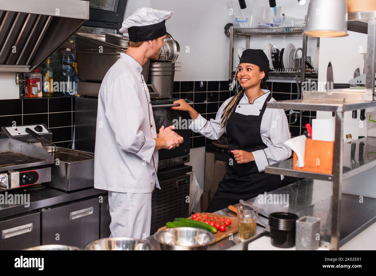 chef in hat and uniform looking at cheerful african american colleague ...