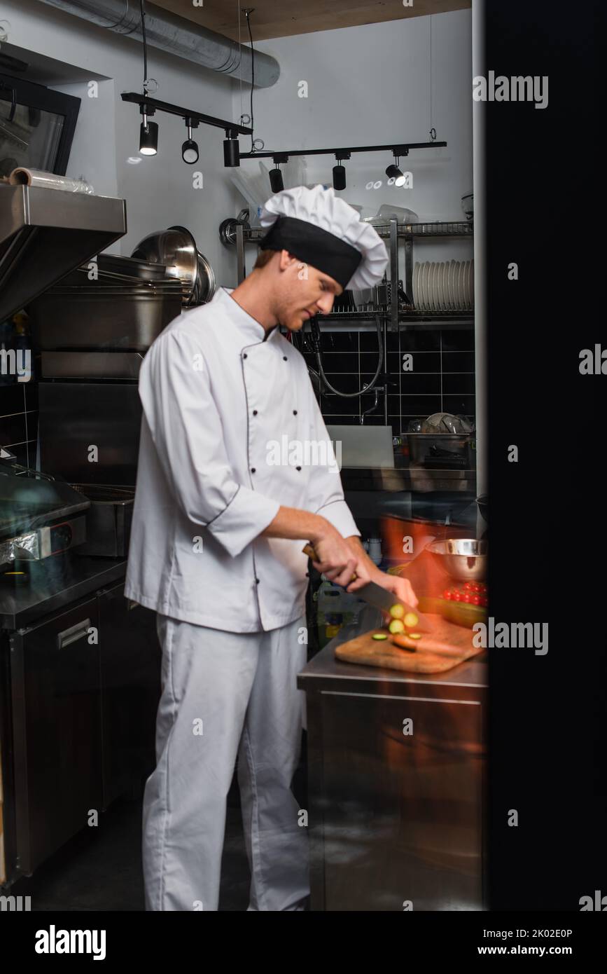young chef in uniform cutting fresh cucumber behind glass door in ...