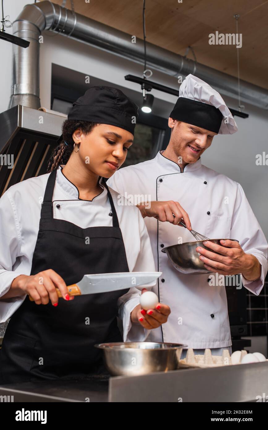 african american sous chef holding knife near raw egg while cooking ...