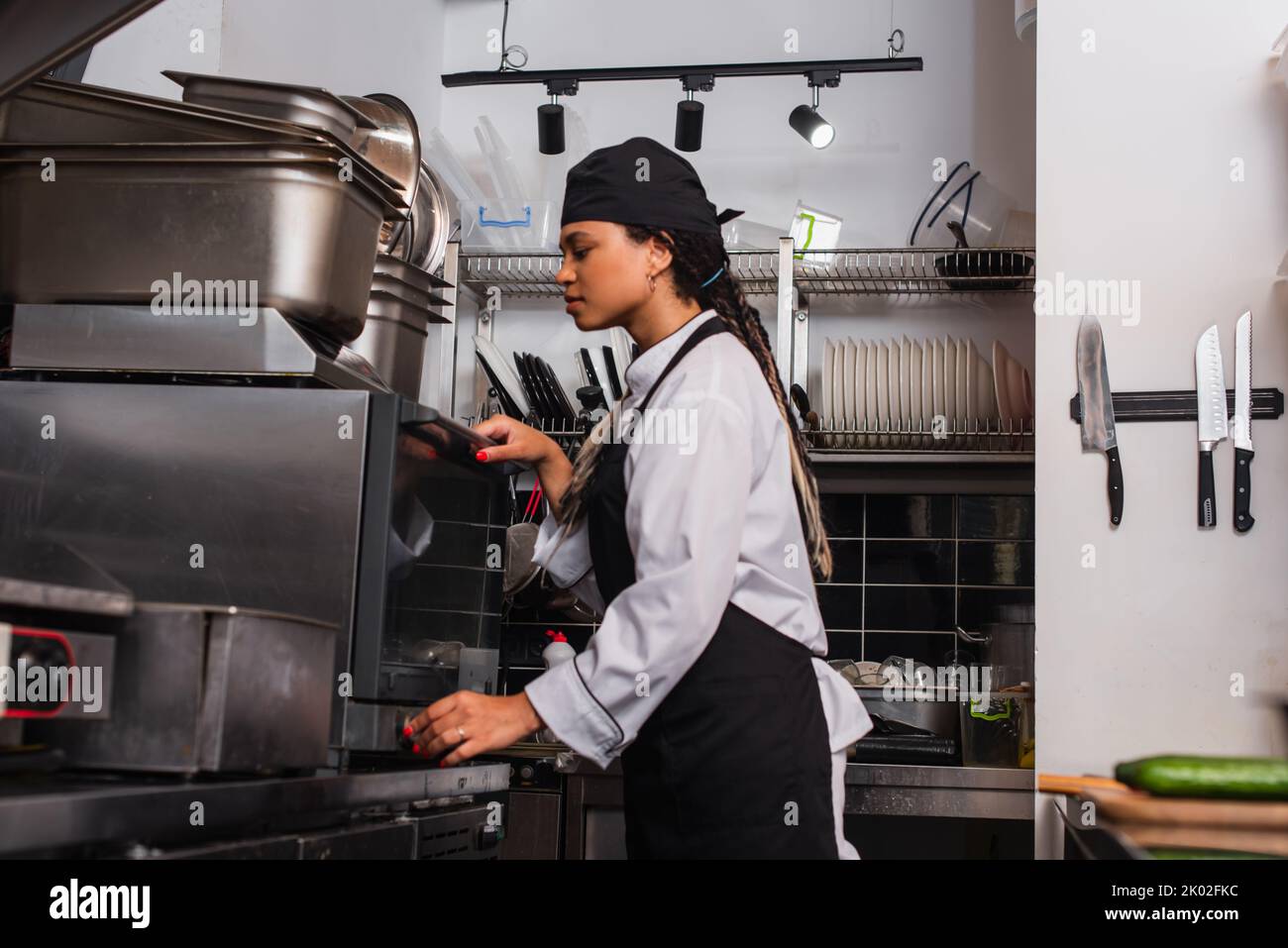 side view of young african american chef in hat using convection oven ...