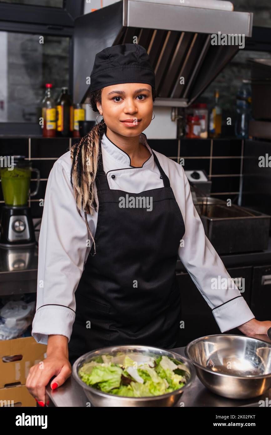 cheerful african american chef in apron standing near bowls and lettuce ...