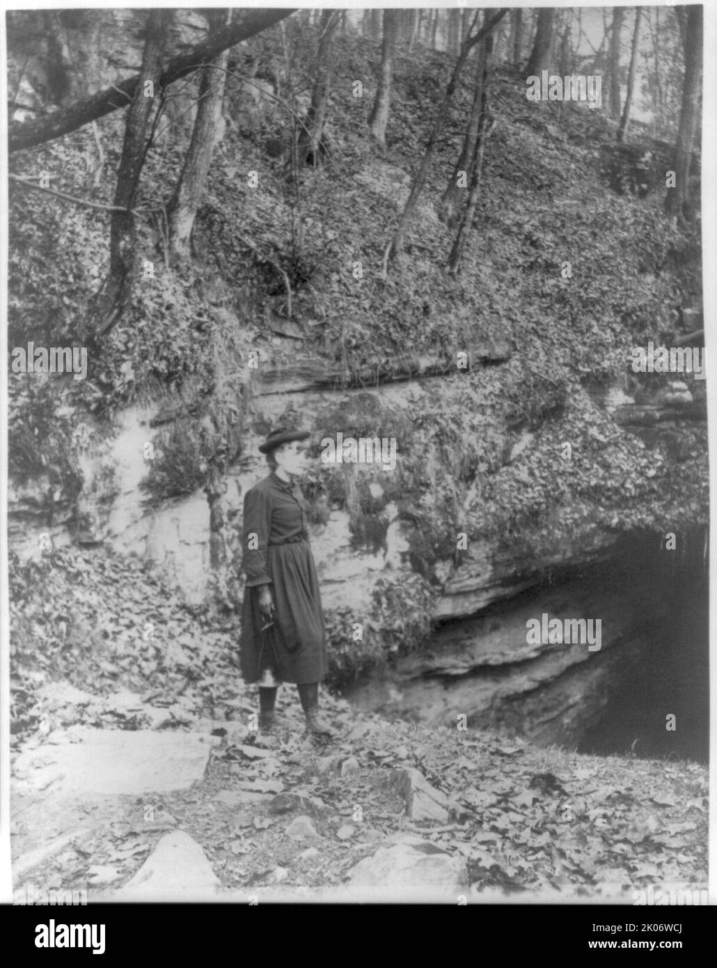 Frances Benjamin Johnston, full, standing by wooded cliff at entrance ...
