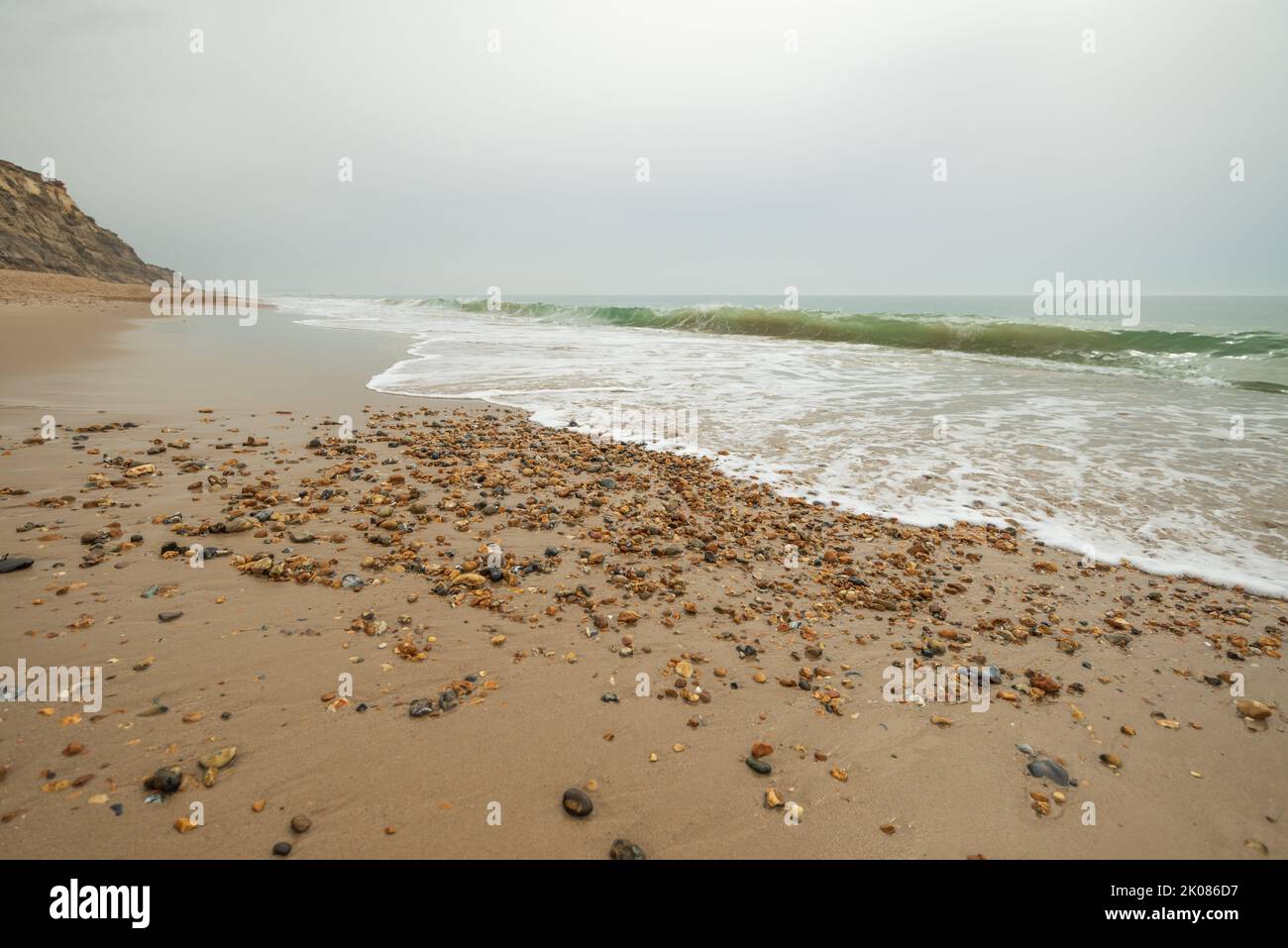 Sandy beach with pebbles and wave washing over, green sea, grey sky ...