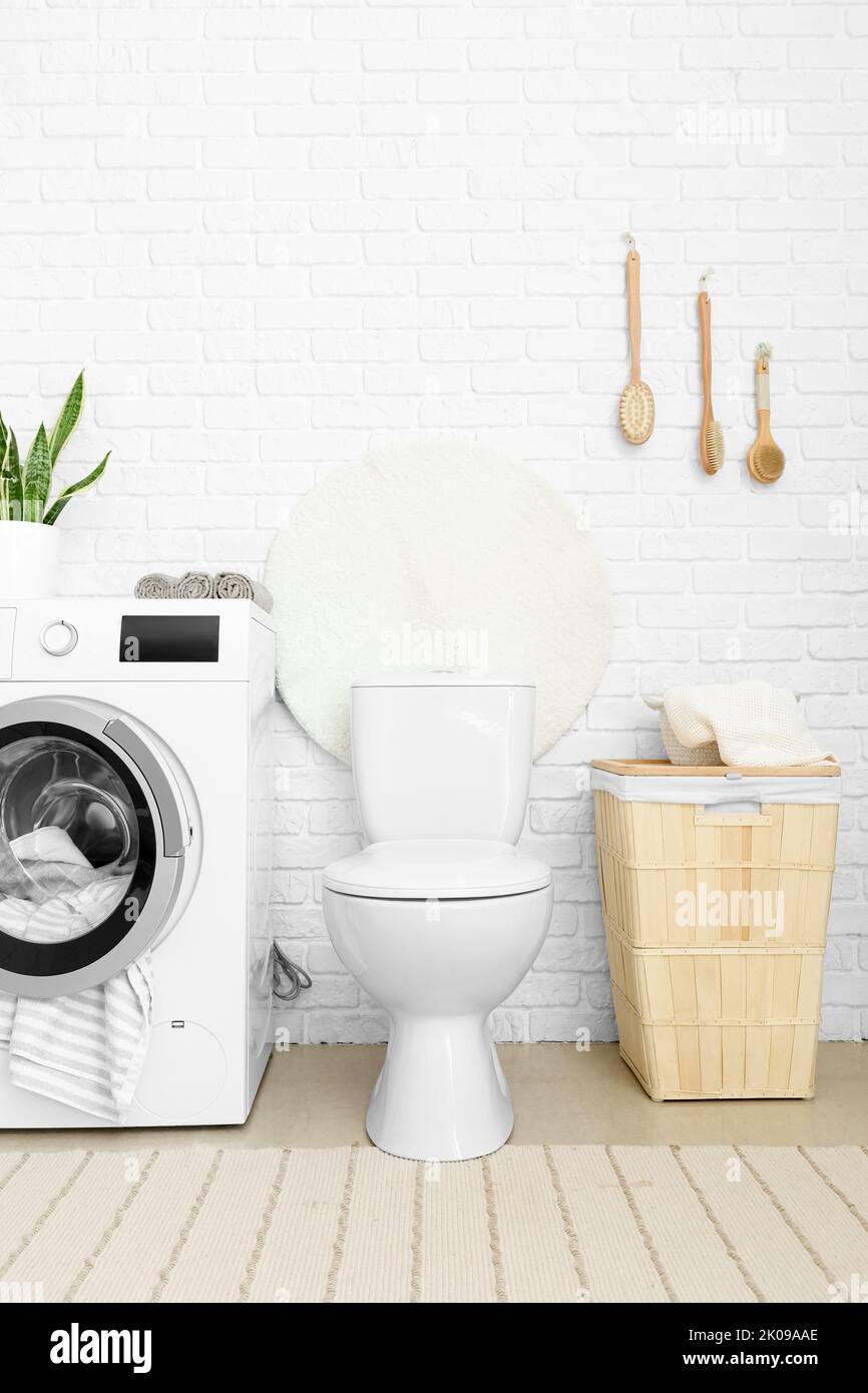 Modern toilet bowl, laundry basket and washing machine near white brick ...
