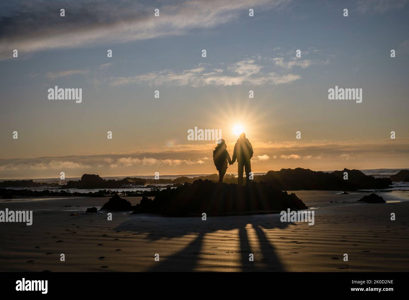 Couple holding hands, watching the sunrise at a rocky beach. Catlins ...