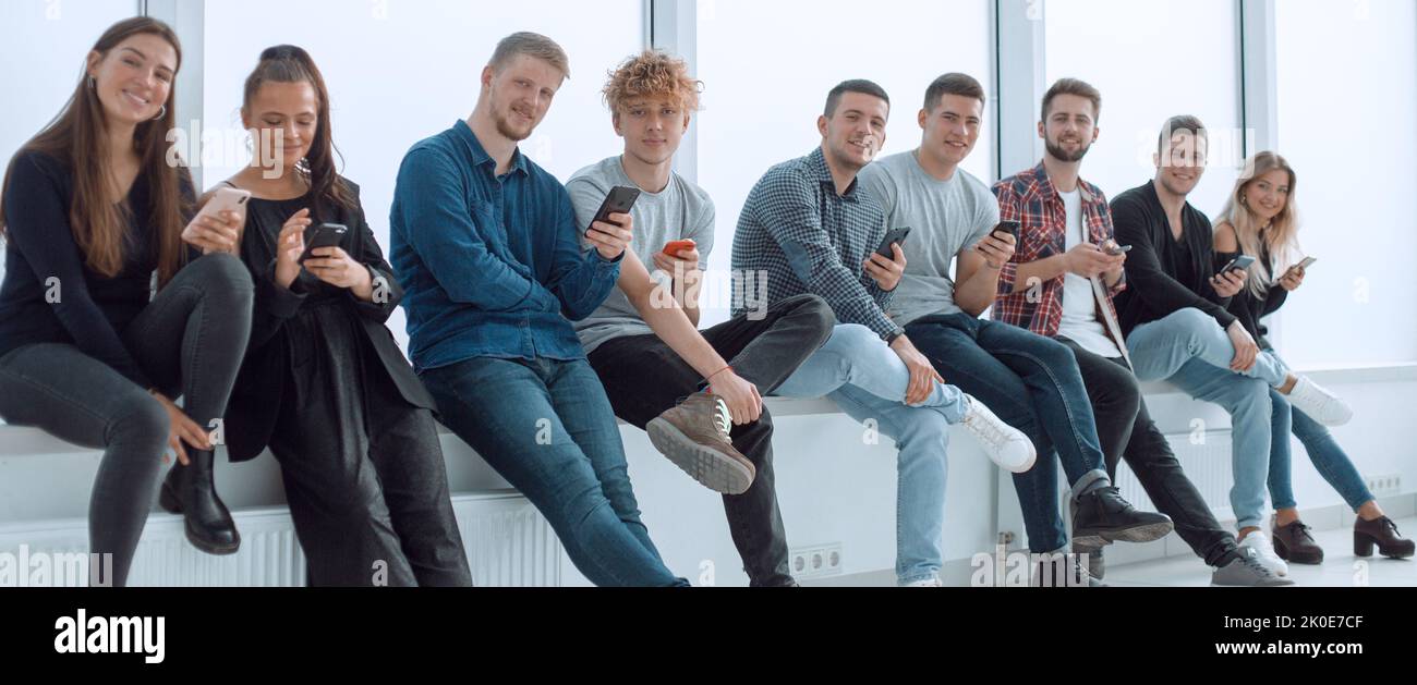 smiling young people with smartphones sitting near an office window ...