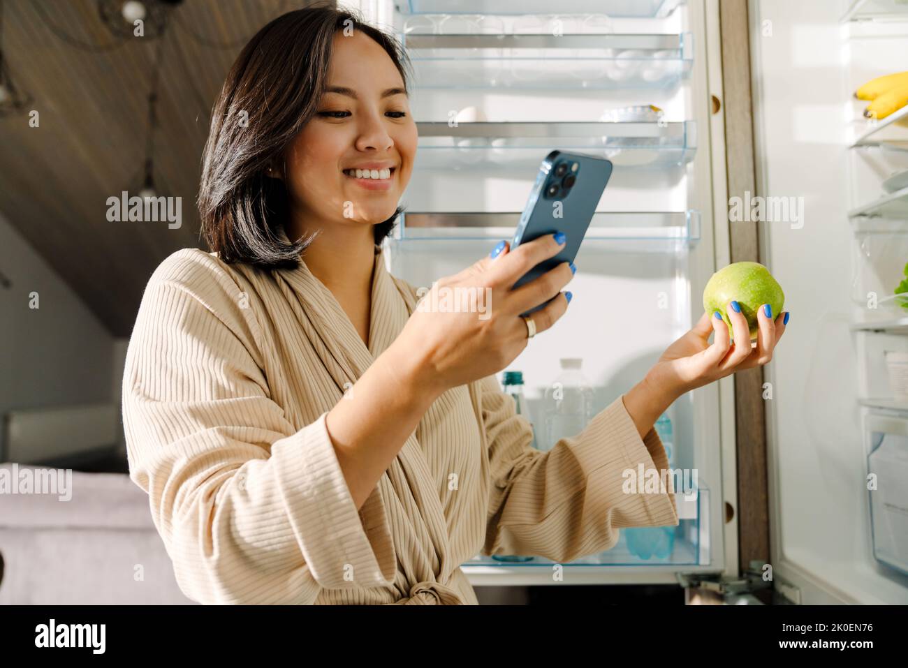 Young asian woman using mobile phone while standing by open fridge at ...