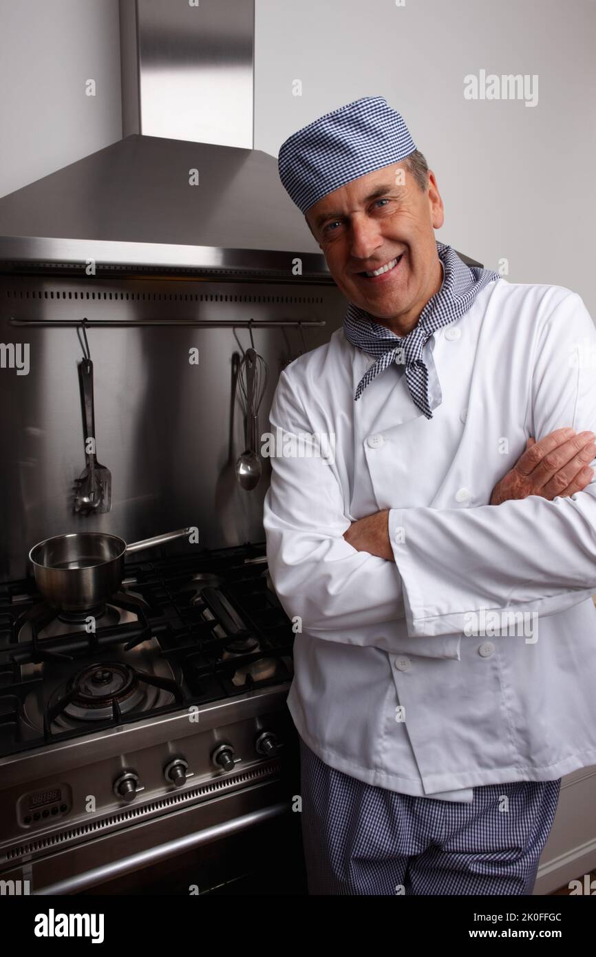 Smiling senior chef with hands folded standing by gas stove. Portrait ...