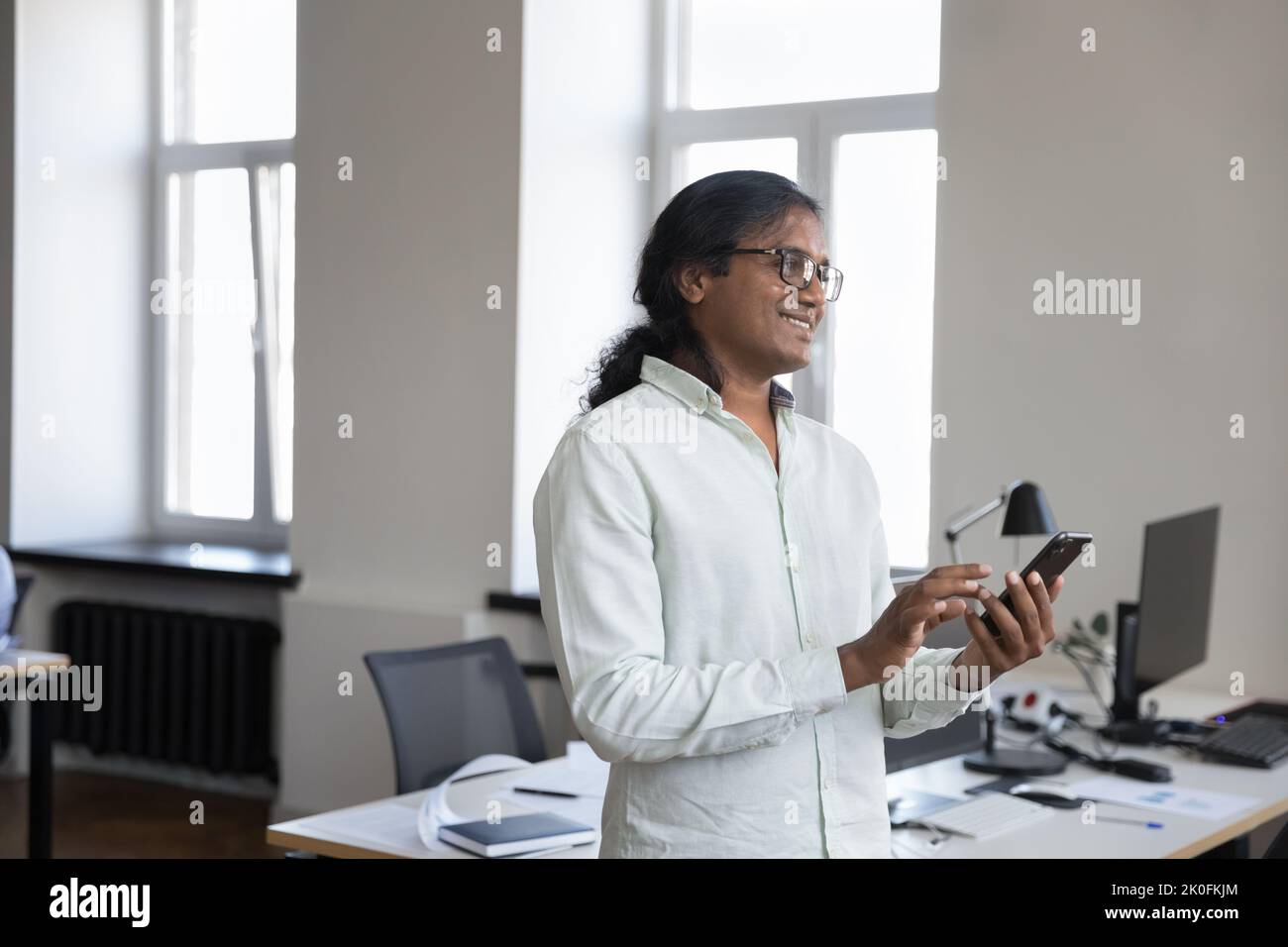 Happy thoughtful Indian employee man holding smartphone, standing at ...