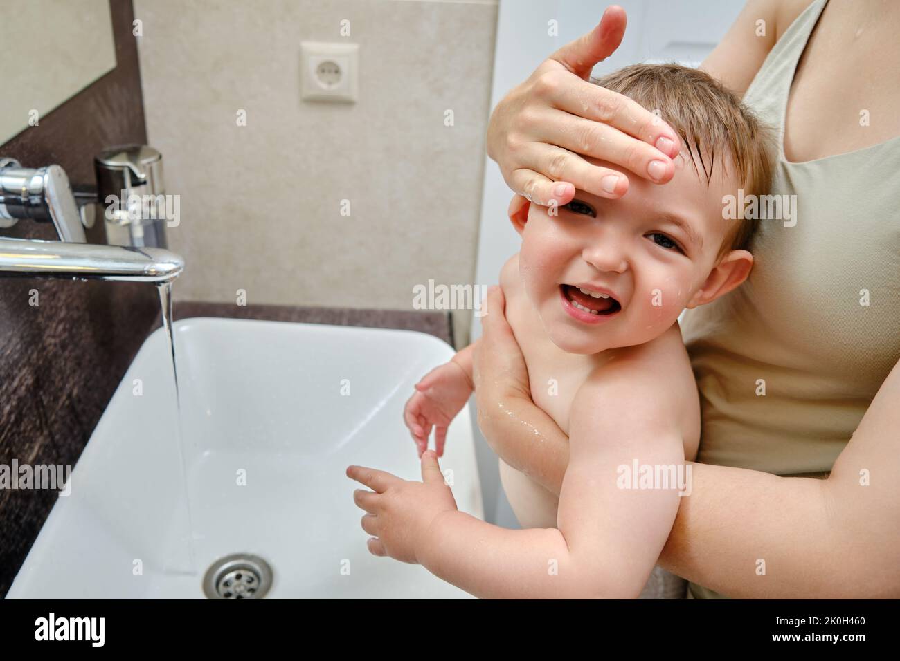 Woman mother washes face of sad toddler baby in sink with faucet. Mom ...