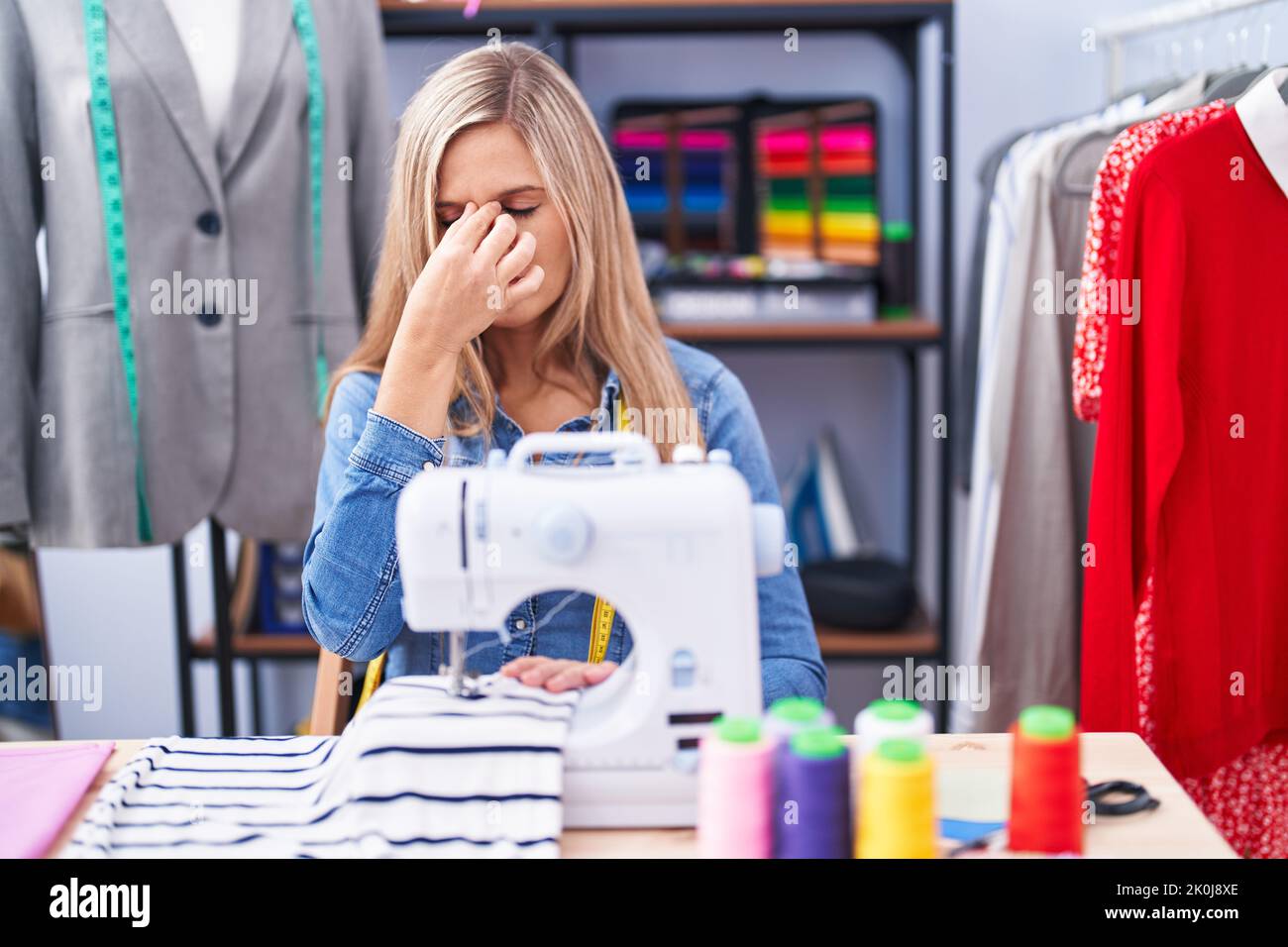 Blonde woman dressmaker designer using sew machine tired rubbing nose ...