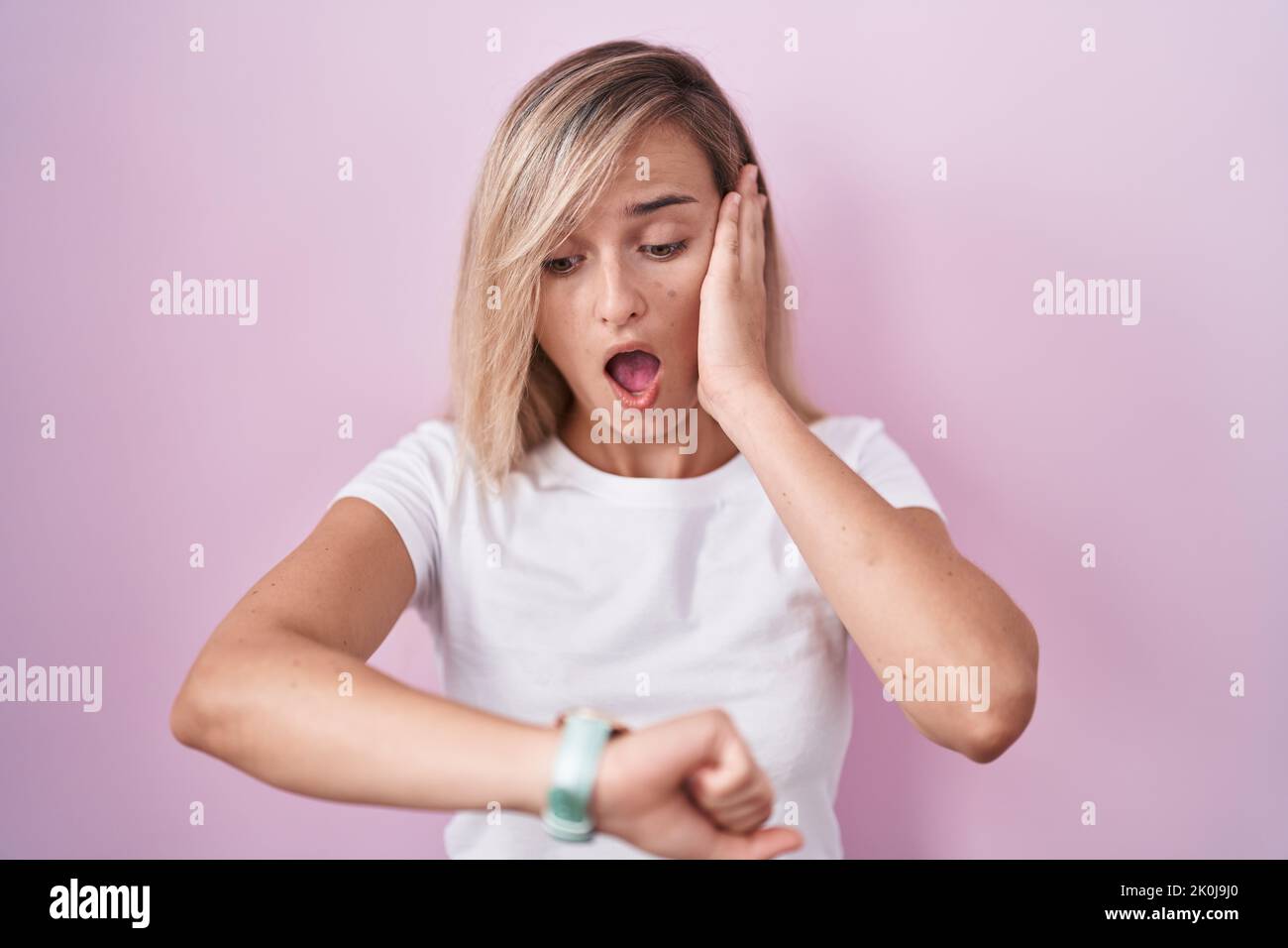 Young blonde woman standing over pink background looking at the watch ...