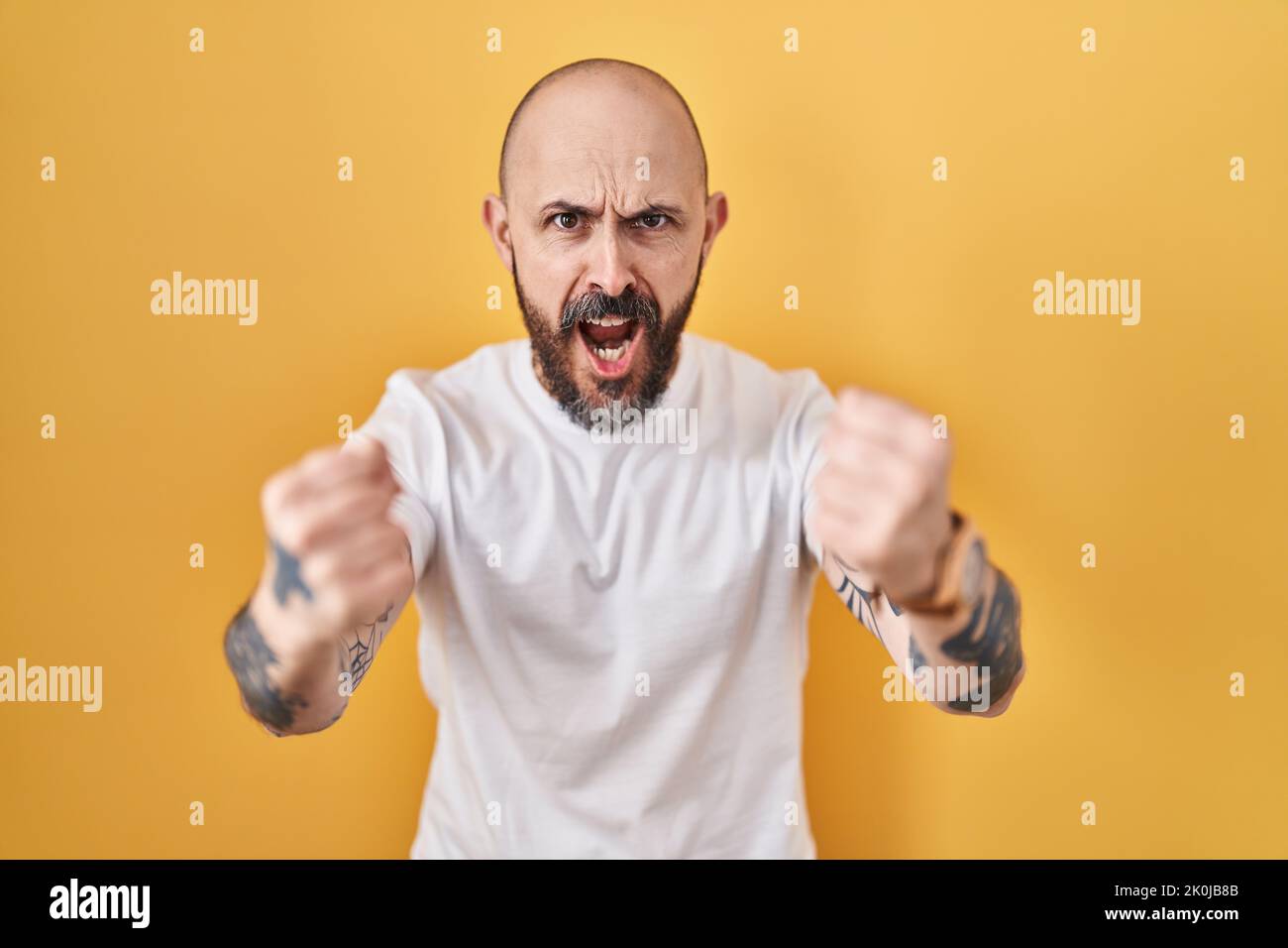 Young hispanic man with tattoos standing over yellow background angry ...
