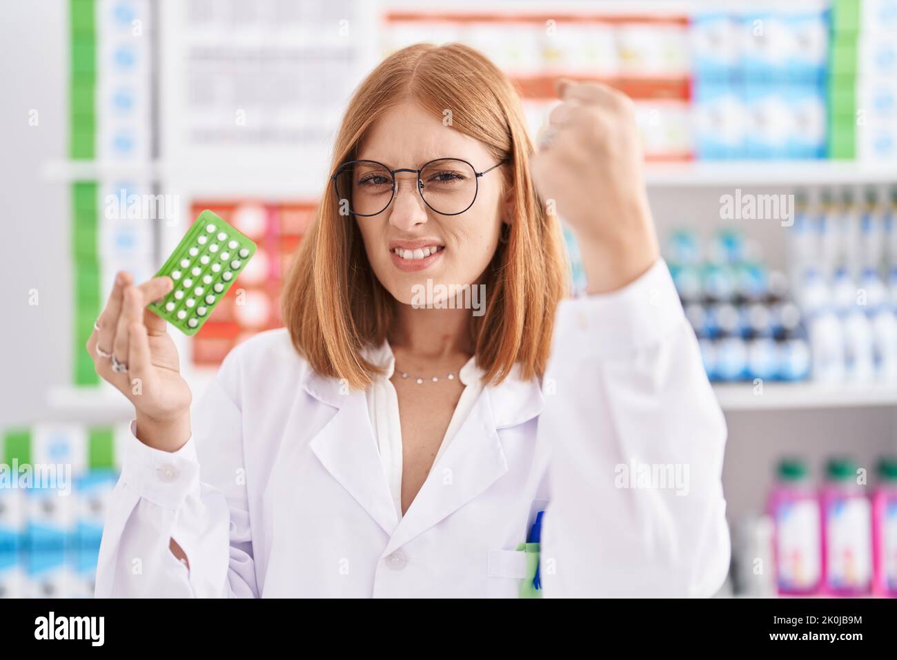Young redhead woman working at pharmacy drugstore holding birth control ...