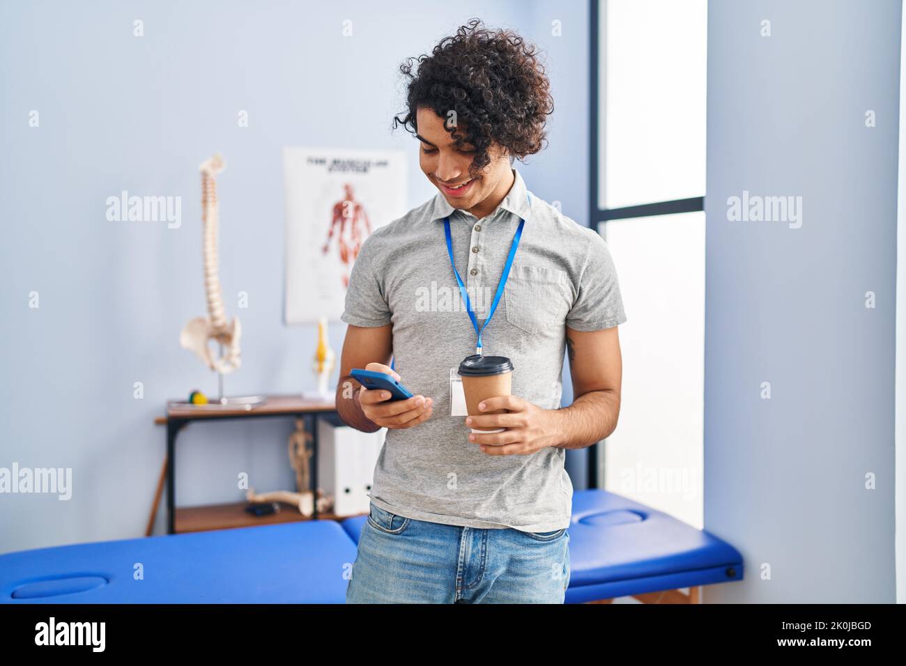 Young hispanic man physiotherapist using smartphone and drinking coffee ...