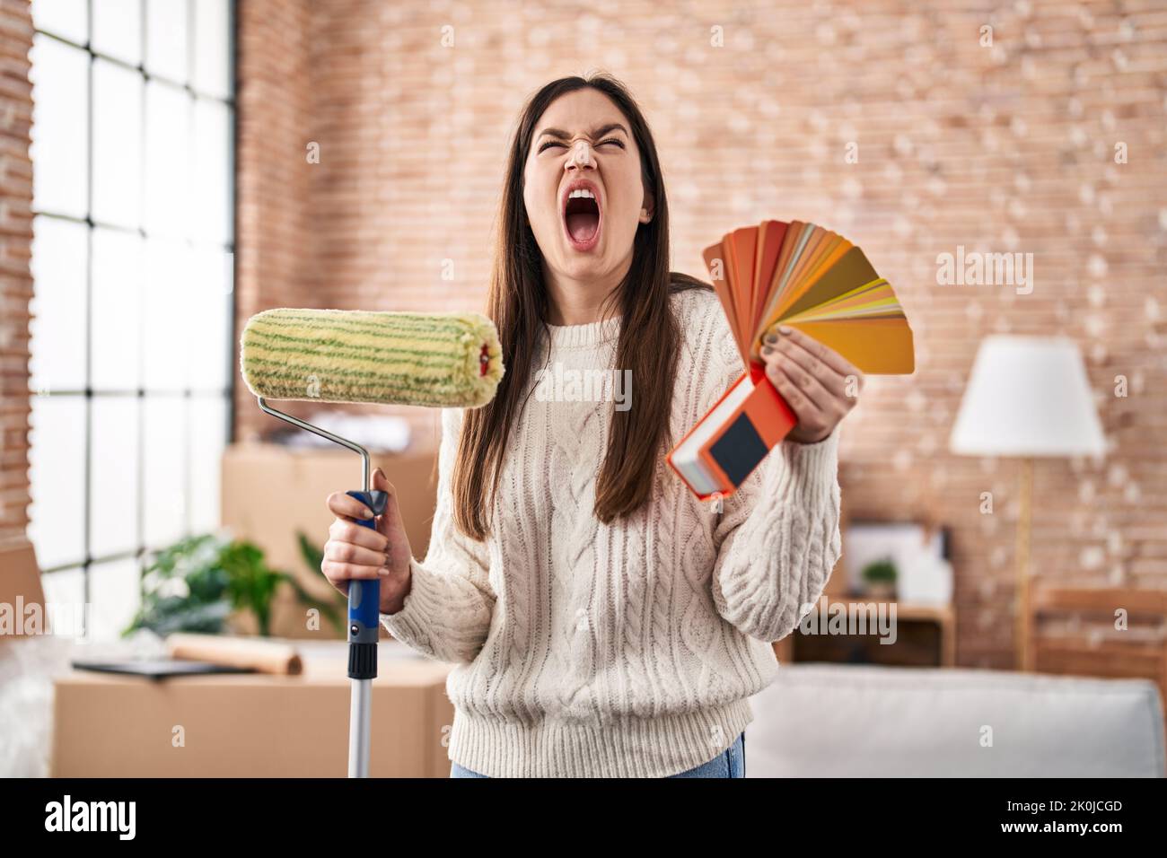 Young brunette woman holding roller painter and paint samples angry and ...