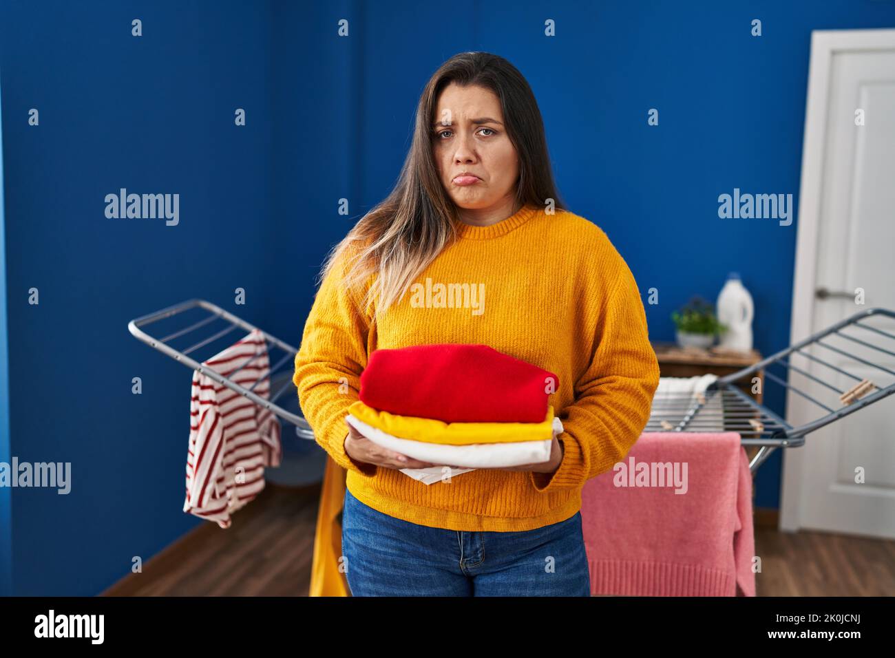 Young hispanic woman holding clean and folded laundry depressed and worry for distress, crying ...