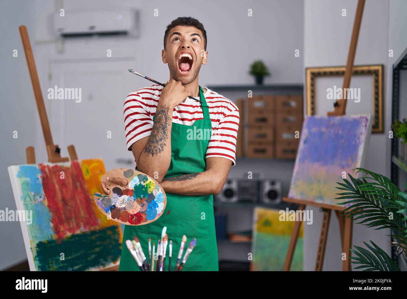 Brazilian young man holding painter palette at artist studio angry and ...