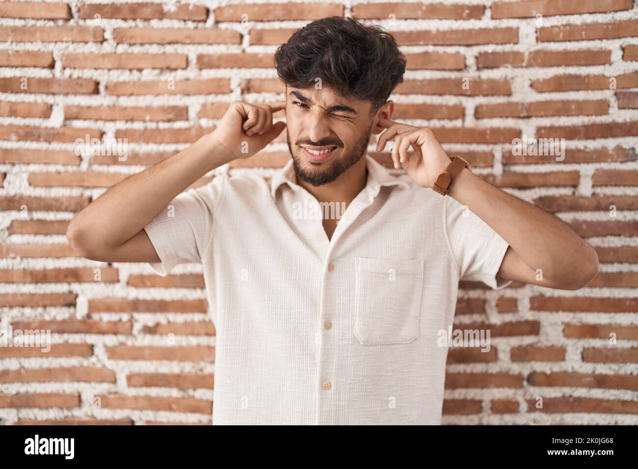 Arab man with beard standing over bricks wall background covering ears ...