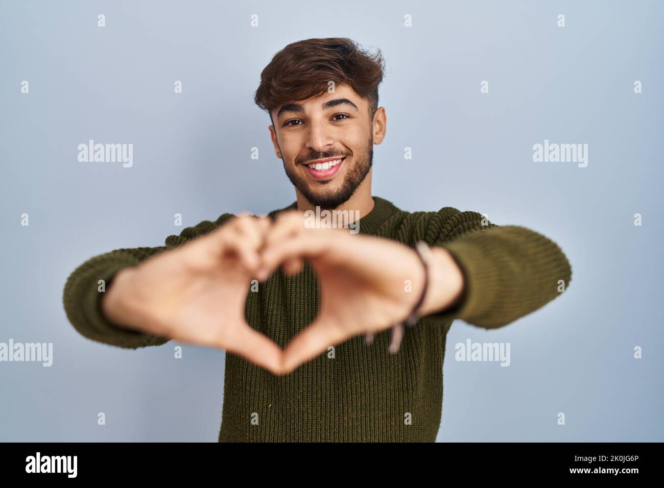 Arab man with beard standing over blue background smiling in love doing ...