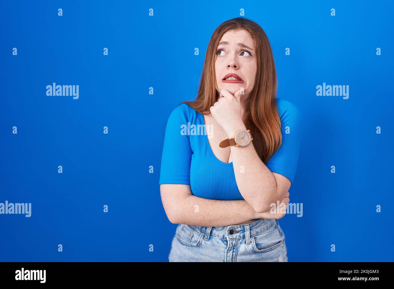 Redhead woman standing over blue background thinking worried about a ...