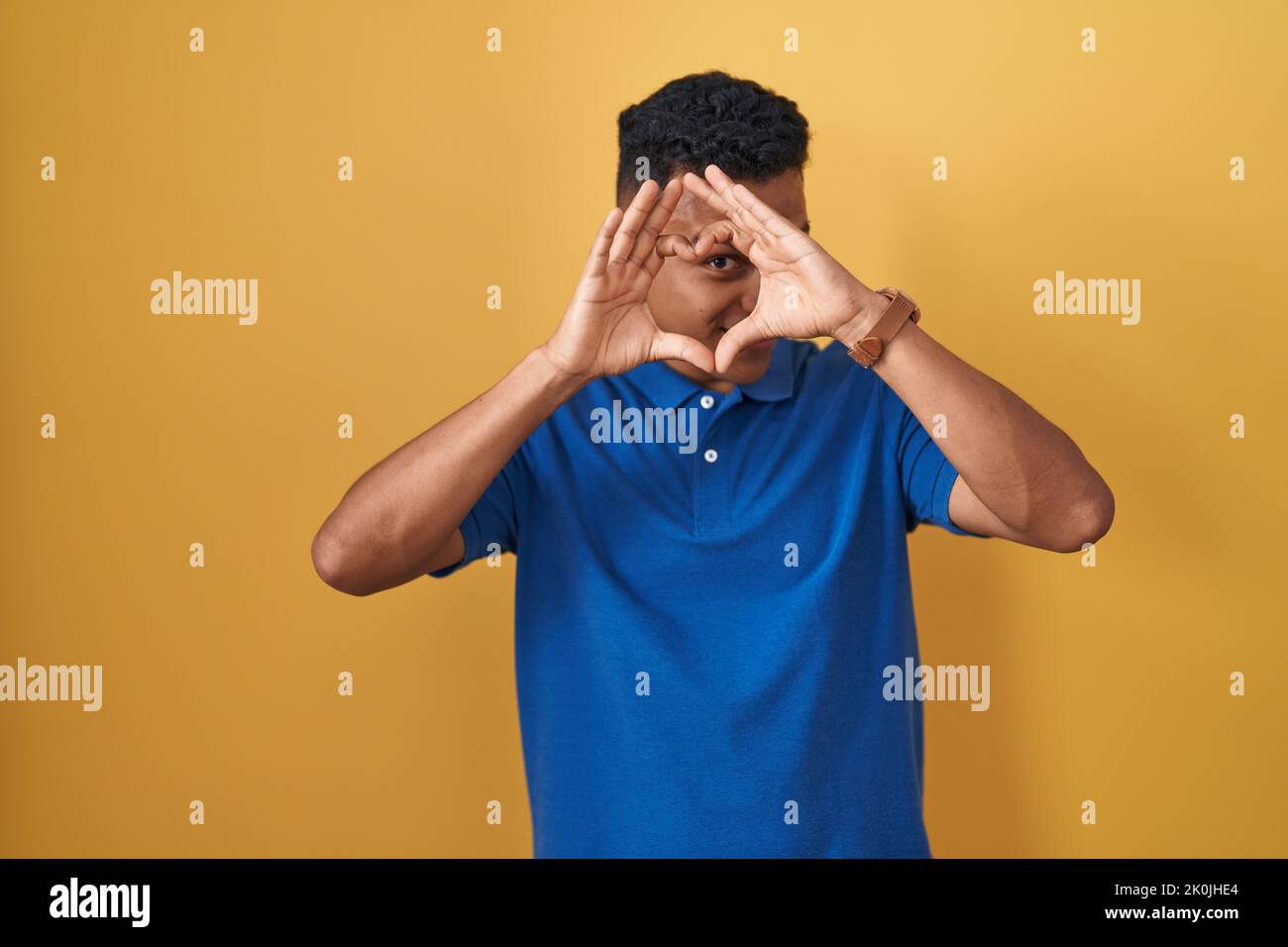 Young hispanic man standing over yellow background doing heart shape ...