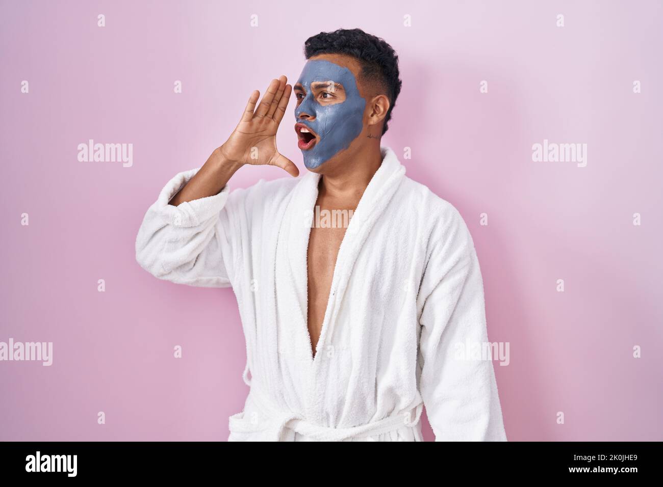 Young hispanic man wearing beauty face mask and bath robe shouting and ...