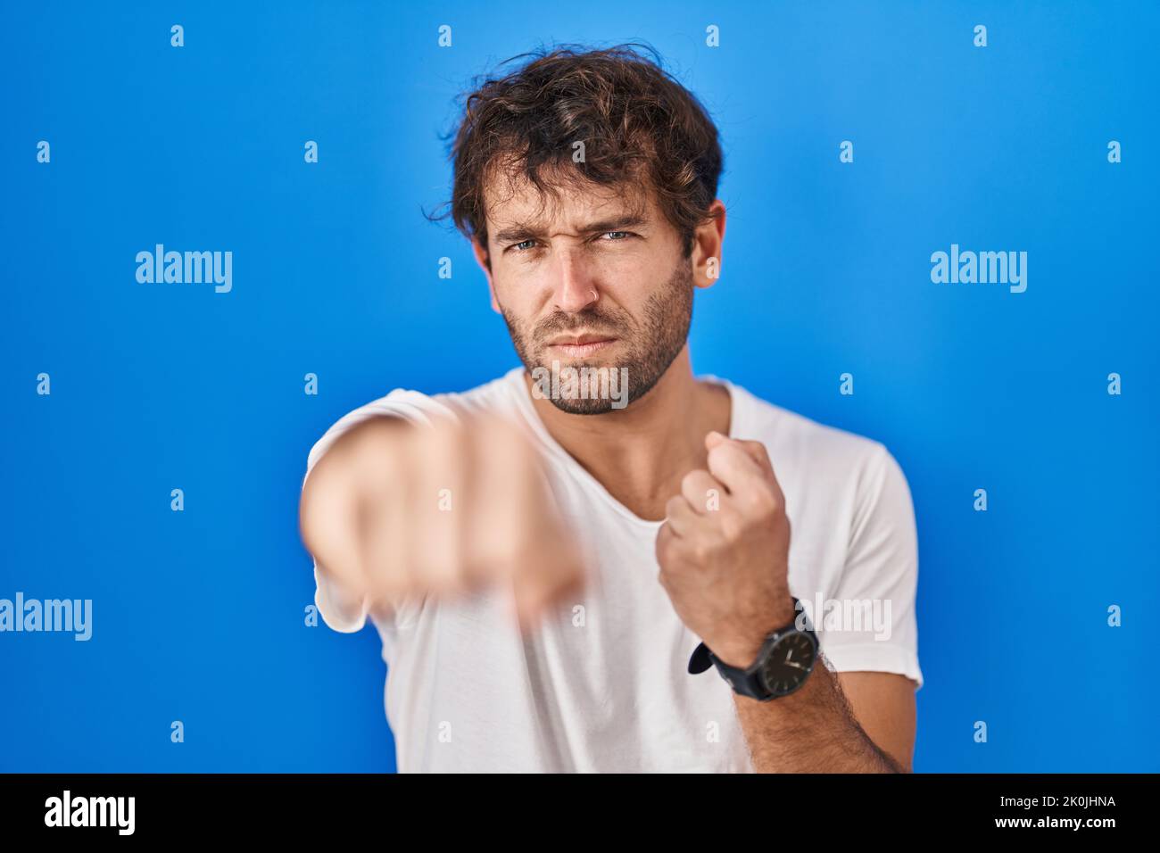 Hispanic young man standing over blue background punching fist to fight ...