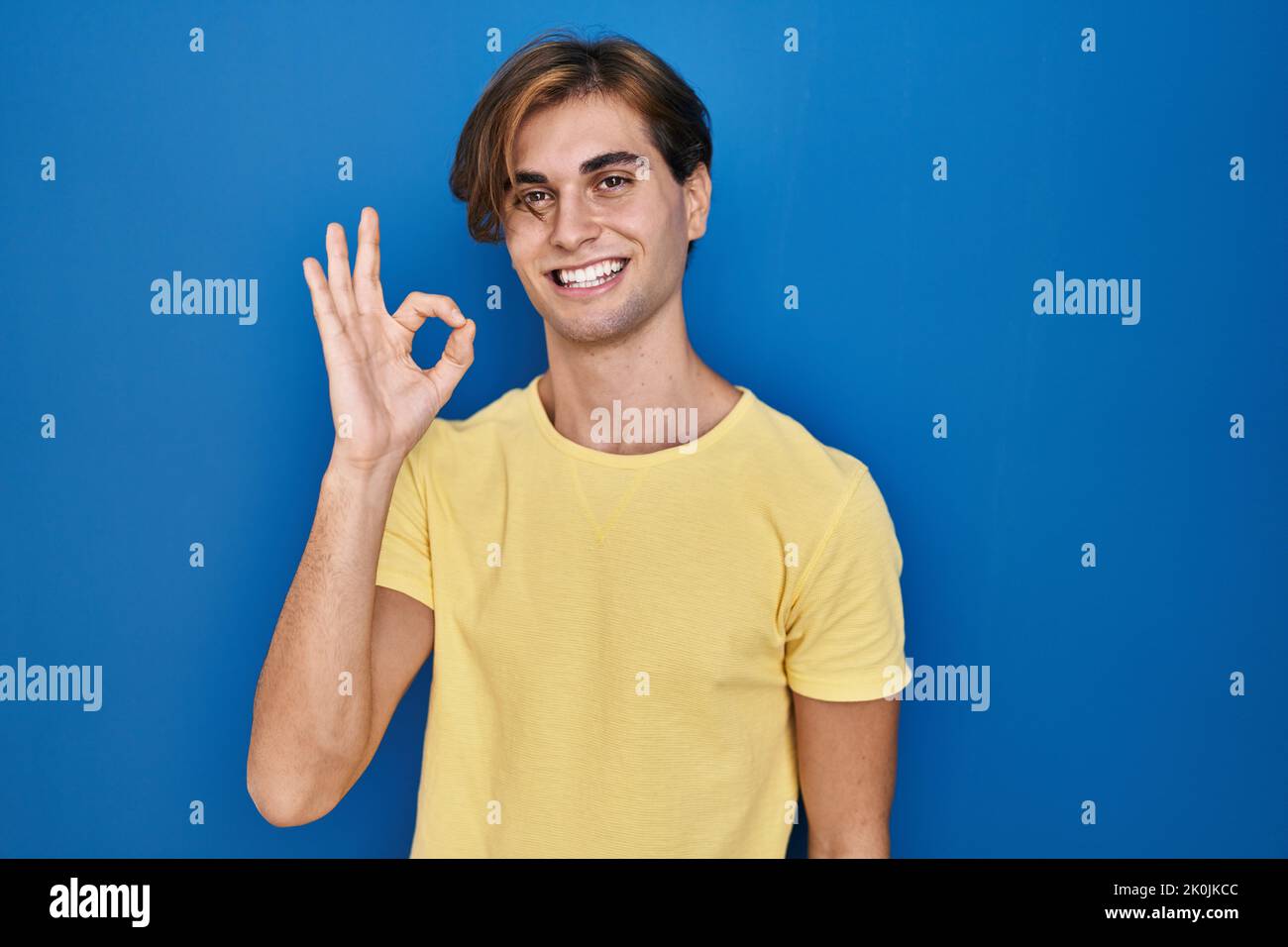 Young man standing over blue background smiling positive doing ok sign ...