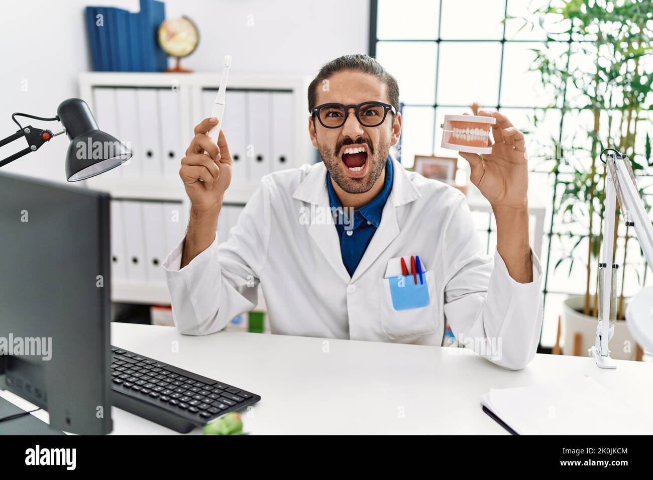 Young hispanic dentist man holding denture and toothbrush angry and mad ...